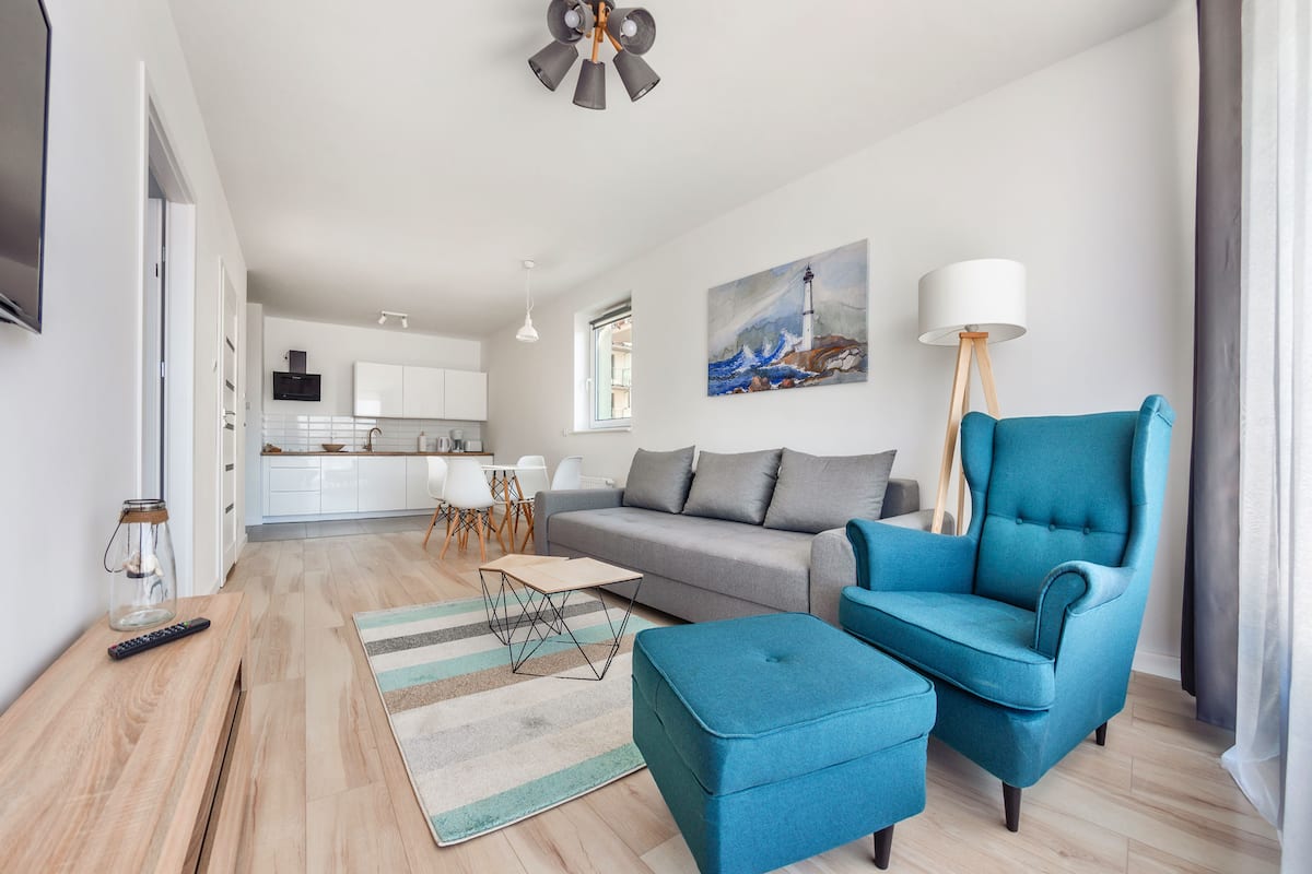 Uma sala de estar moderna com um sofá, um banquinho e uma poltrona azul. Há um quadro na parede, um abajur e um controle remoto em uma mesa de madeira.