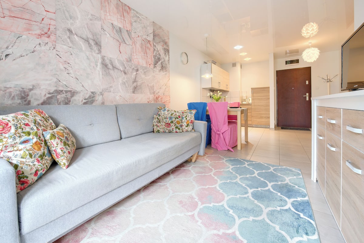 Uma sala de estar com sofá, almofadas florais, parede de mármore, área de jantar com mesa e cadeiras, e um guarda-roupa.
