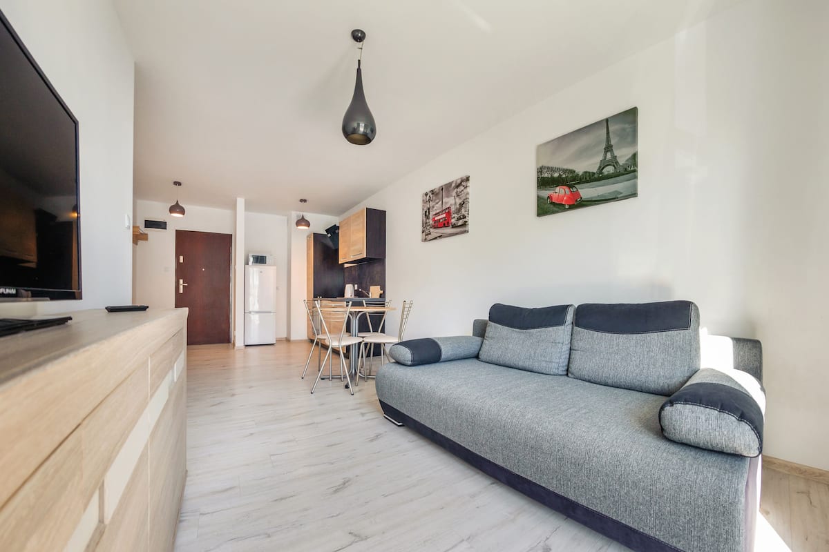 Uma sala de estar moderna com um sofá cinza, mesa de jantar de madeira e cozinha ao fundo.