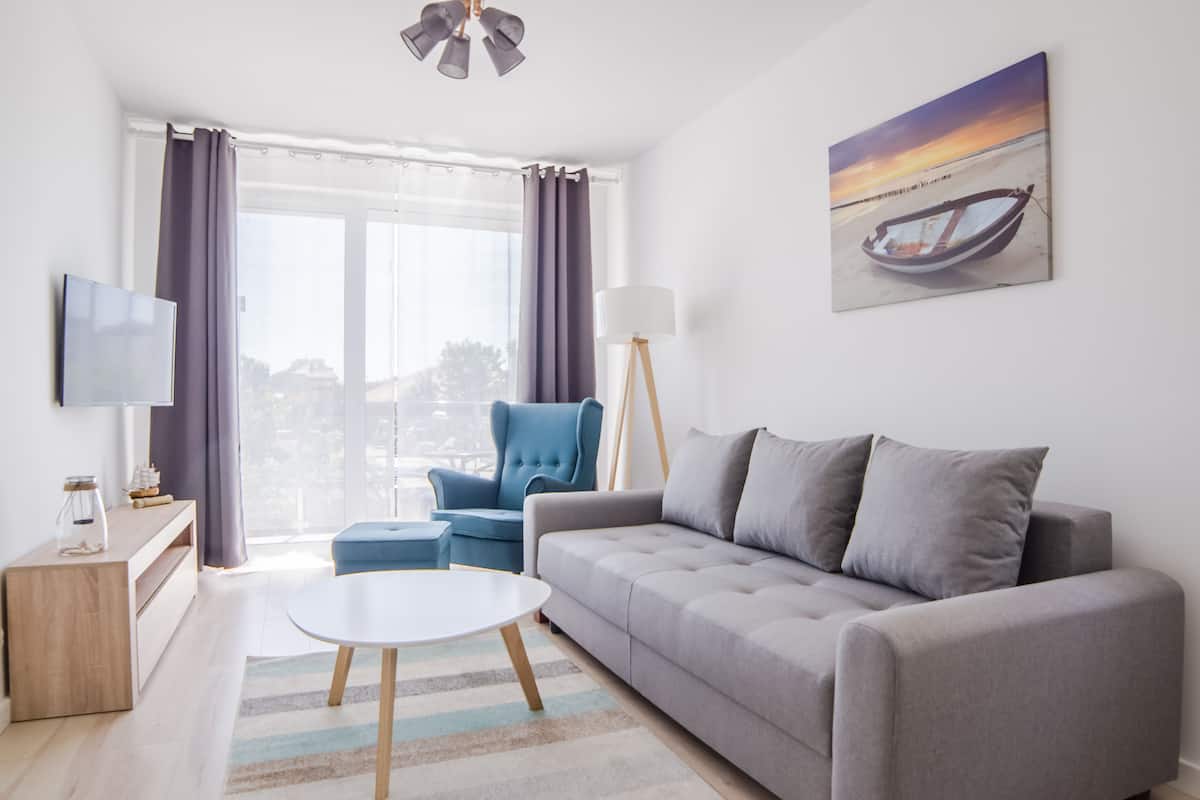 Uma sala de estar moderna com um sofá cinzento, uma poltrona azul, uma mesa de centro branca e um quadro de um barco na praia.