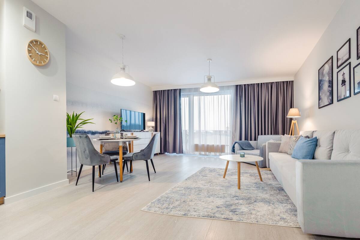 Uma sala de estar moderna com um sofá, uma mesa de jantar com cadeiras, uma televisão e uma janela grande com cortinas.