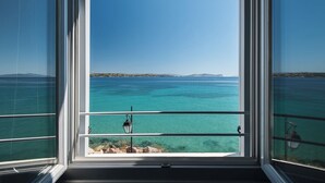 Vista da janela para um mar azul e límpido, com uma linha de costa distante e um candeeiro na balaustrada da varanda.
