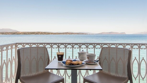 Uma varanda com duas cadeiras, uma mesa com um prato de pastelaria, uma chávena de café e um bule, com vista para um mar calmo e montanhas ao longe.