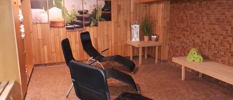 Sala de spa com paredes de madeira, piso ladrilhado e duas cadeiras de massagem pretas. Há uma pequena mesa com um relógio e um vaso de planta.