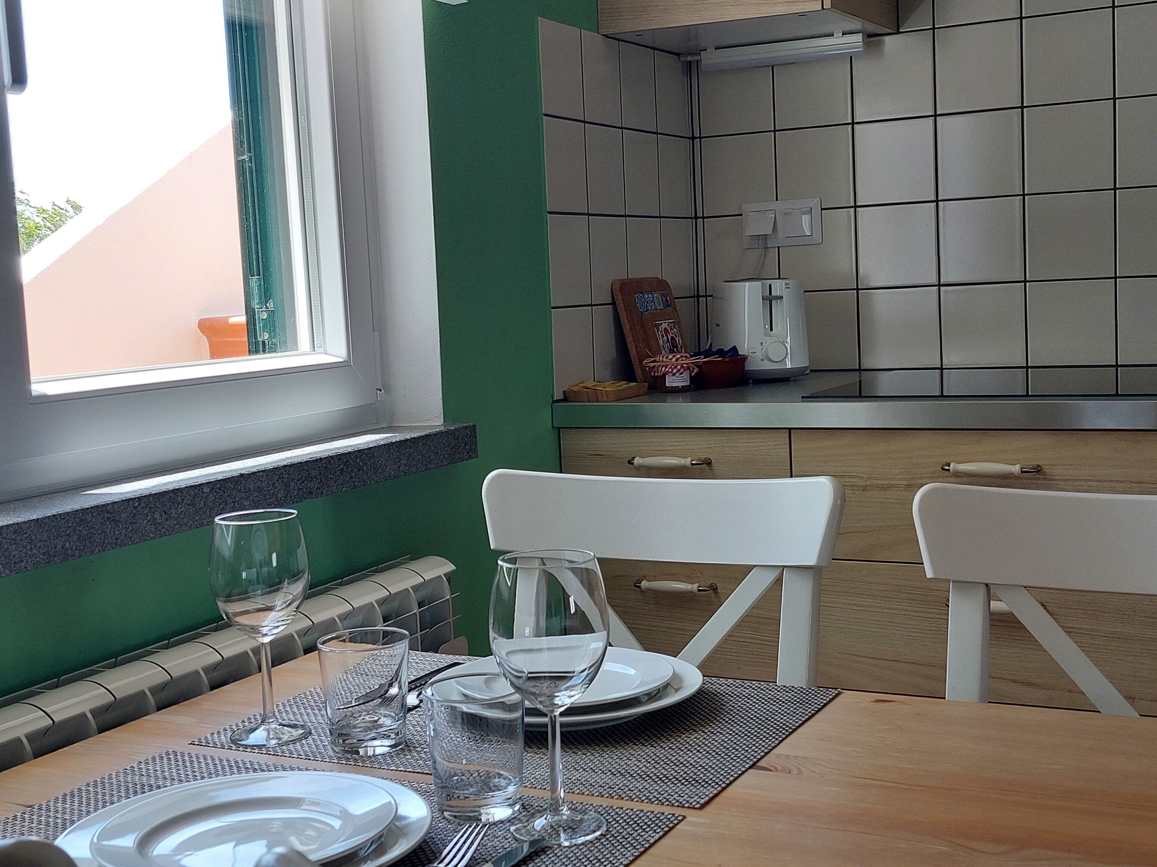 Uma área de refeições com uma mesa posta para dois, uma janela, uma cozinha com painel de azulejos e um balcão com uma torradeira.