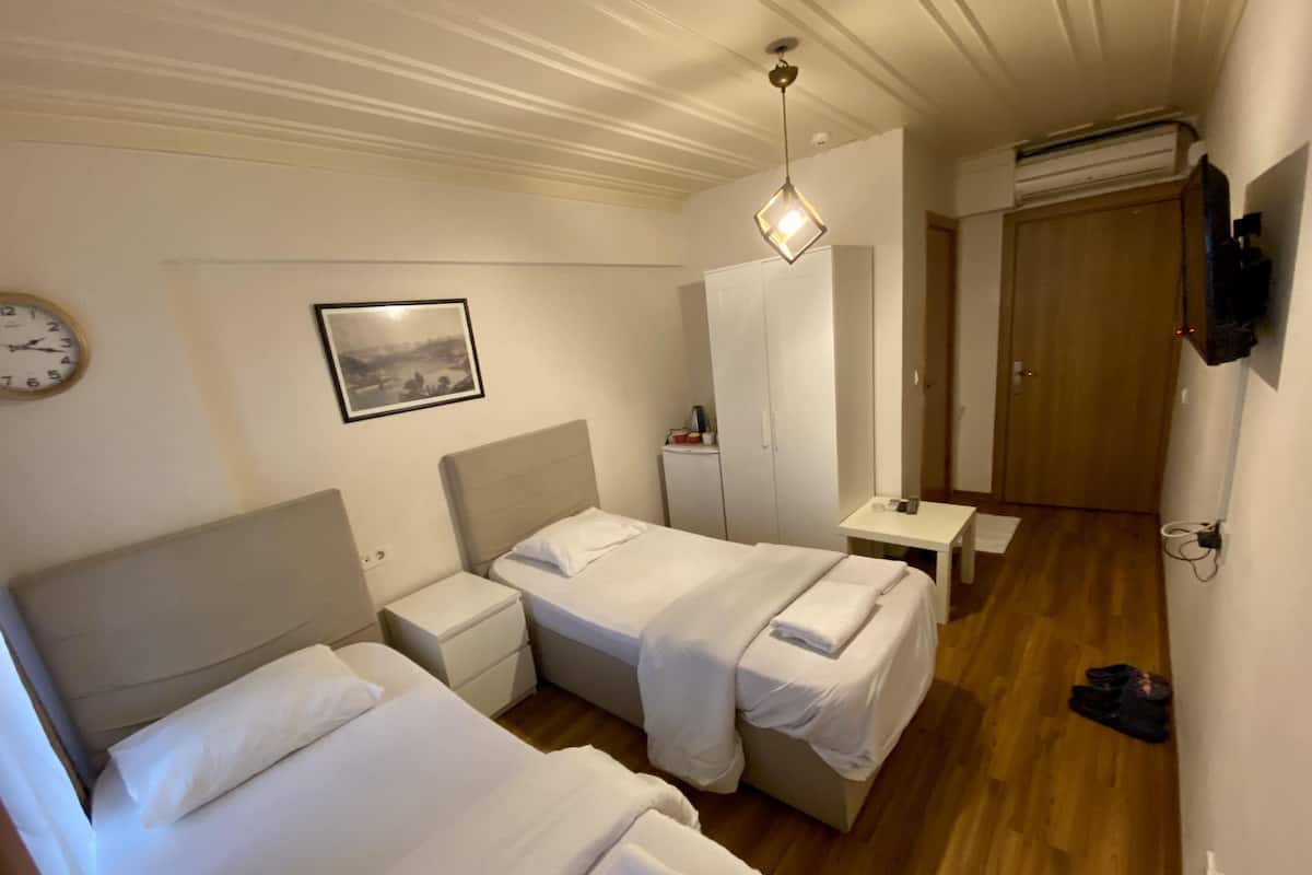 Quarto de hotel com duas camas, uma mesinha de cabeceira, uma escrivaninha, um relógio, um quadro e um aparelho de ar condicionado.