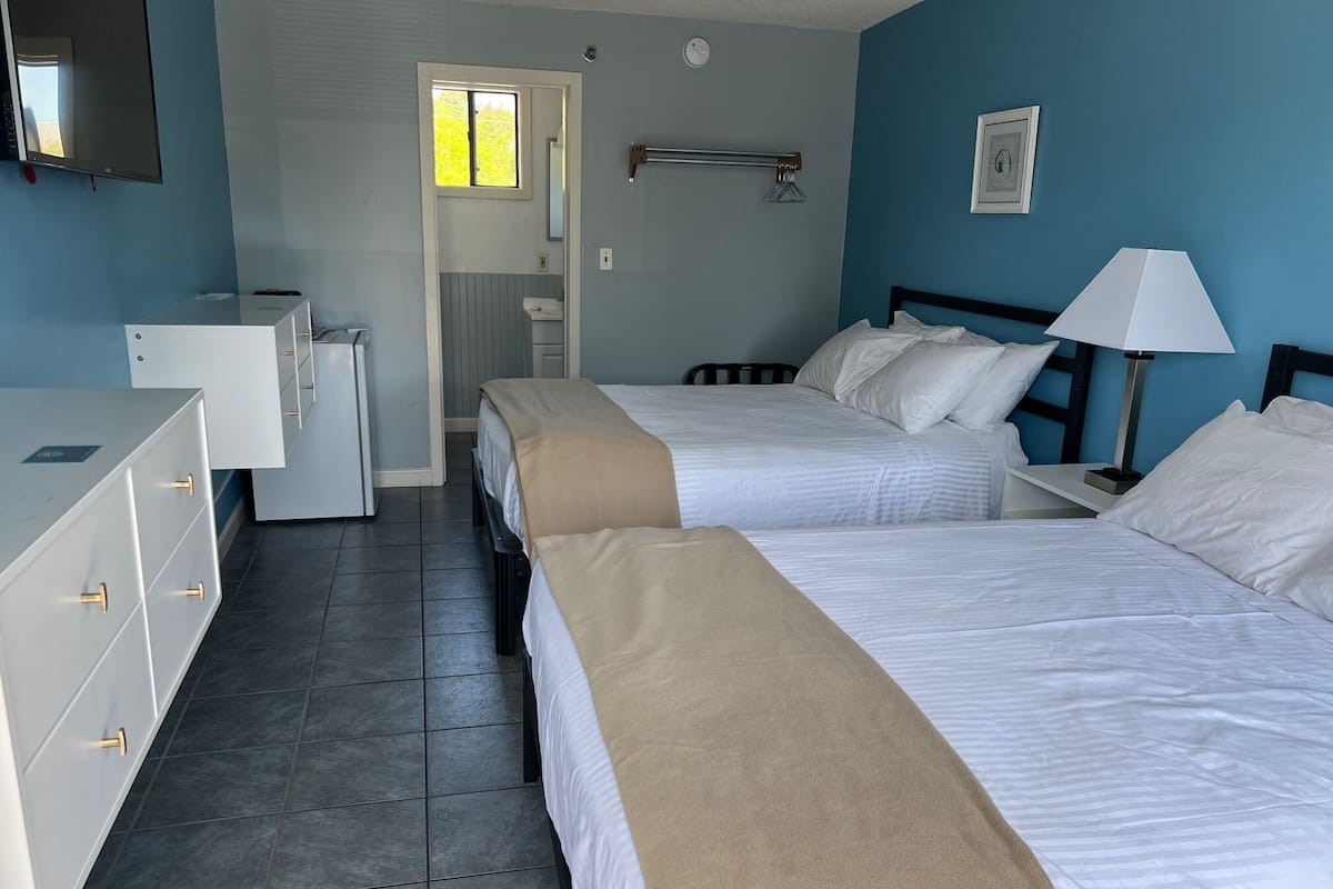 Quarto de hotel com duas camas, uma escrivaninha, uma cadeira, um abajur e uma janela.