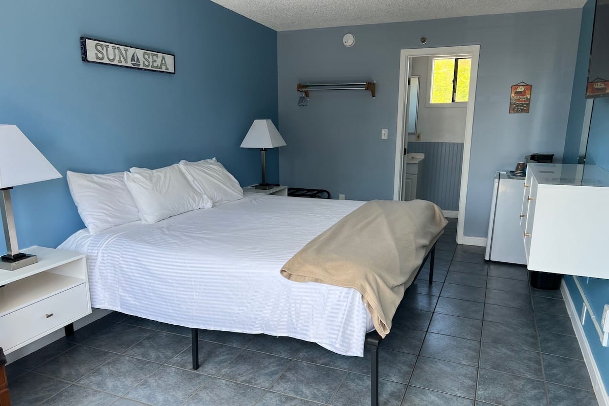 Quarto com cama, mesas de cabeceira, cômoda e porta para o banheiro.