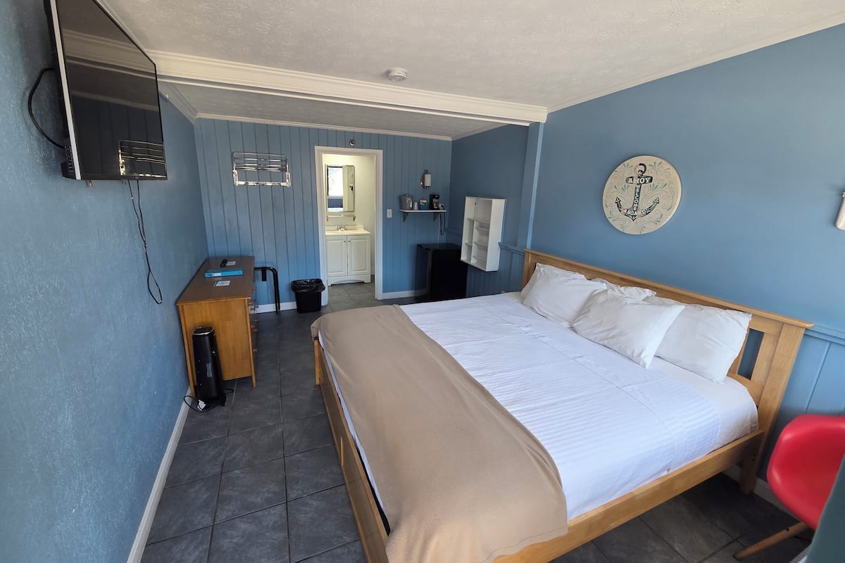 Quarto de hotel com cama, escrivaninha, cadeira, televisão fixada na parede e banheiro visível através de uma porta aberta.