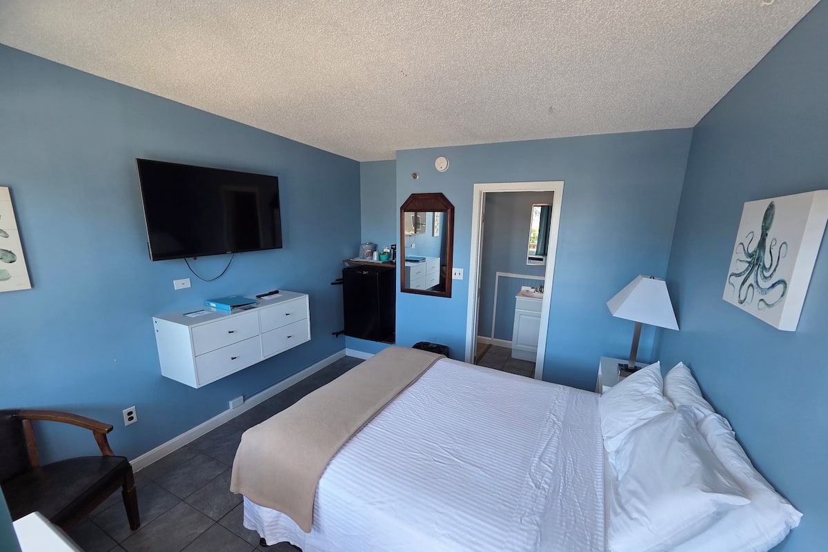 Quarto com cama, escrivaninha, cadeira, televisão, cômoda, espelho e banheiro visível através de uma porta aberta.