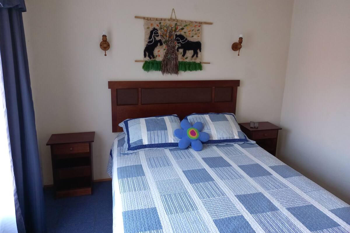 Uma cama bem arrumada com colcha estampada, duas mesas de cabeceira de madeira, uma tapeçaria na parede e um cabeceiro de madeira.