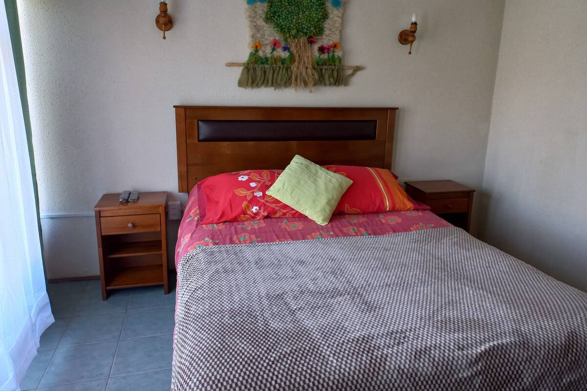 Uma cama bem arrumada com colcha estampada, cabeceira de madeira e duas mesas de cabeceira com abajures.