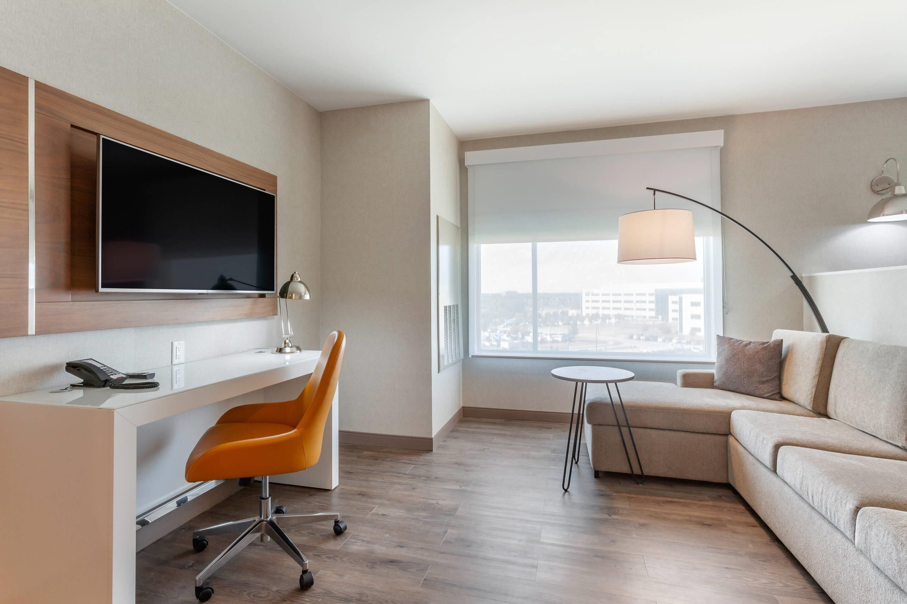 Quarto de hotel moderno com televisão de tela plana, escrivaninha com cadeira, sofá, mesa redonda pequena e uma grande janela com vista para a cidade.