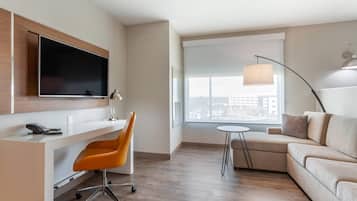 Quarto de hotel moderno com televisão de tela plana, escrivaninha com cadeira, sofá, mesa redonda pequena e uma grande janela com vista para a cidade.