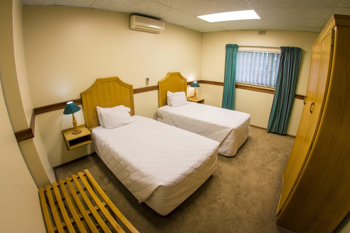 Quarto de hotel com duas camas de solteiro, cabeceiras de madeira, mesas de cabeceira com luminárias, uma janela com cortinas e um guarda-roupa.