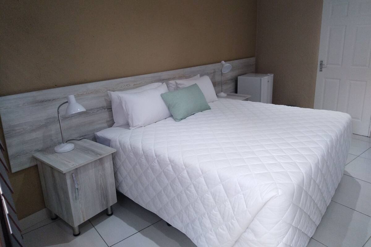 Uma cama bem arrumada com um espalha branco acolchoado, cabeceira de madeira e mesa de cabeceira com abajur.
