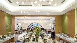 Uma ampla área de buffet com vários pontos de serviço, um arranjo floral central e uma grande janela em arco ao fundo.