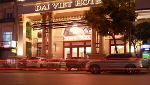 Entrada do Hotel Dai Viet com uma placa, uma árvore grande e carros estacionados.