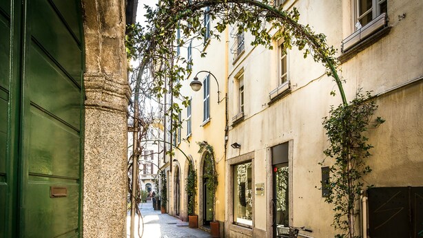 Uma viela estreita com uma porta verde à esquerda, uma bicicleta vermelha à direita e edifícios com janelas em arco e plantas em vasos.