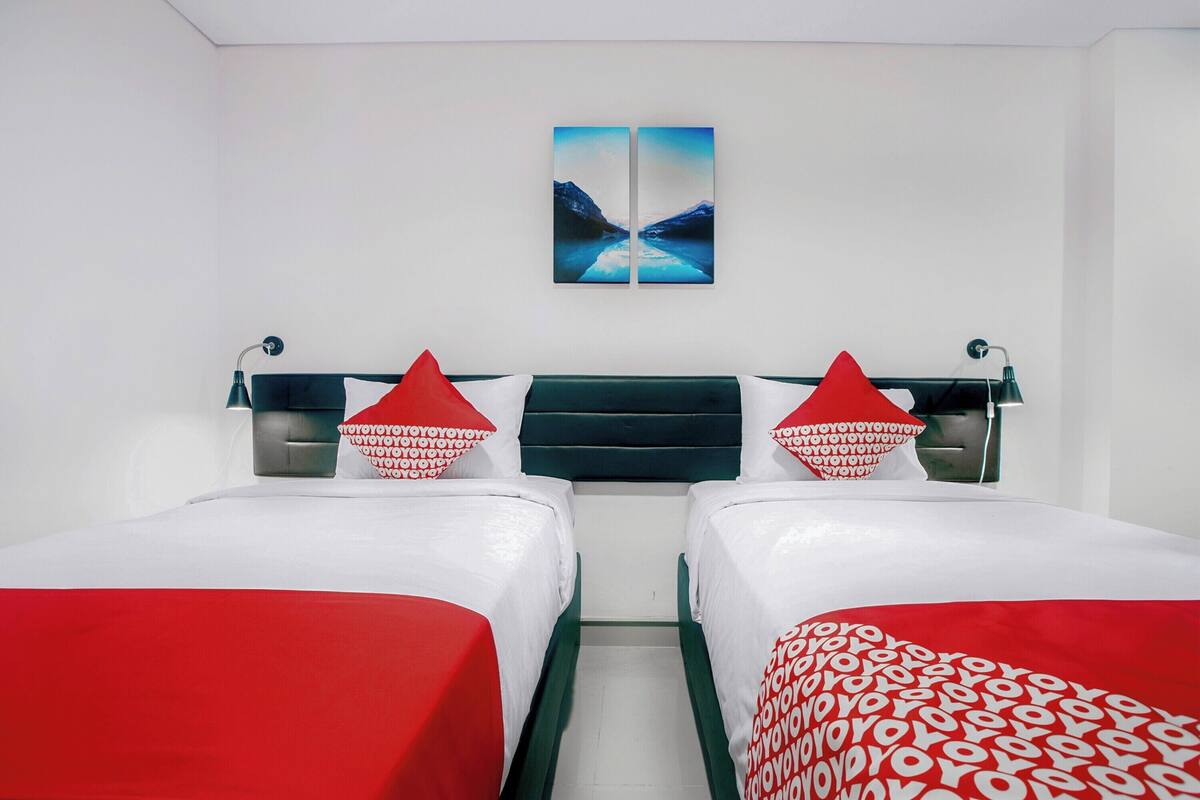 Quarto de hotel com duas camas, cada uma com colcha vermelha e branca, uma obra de arte azul e branca acima das camas e cabeceiras verdes.