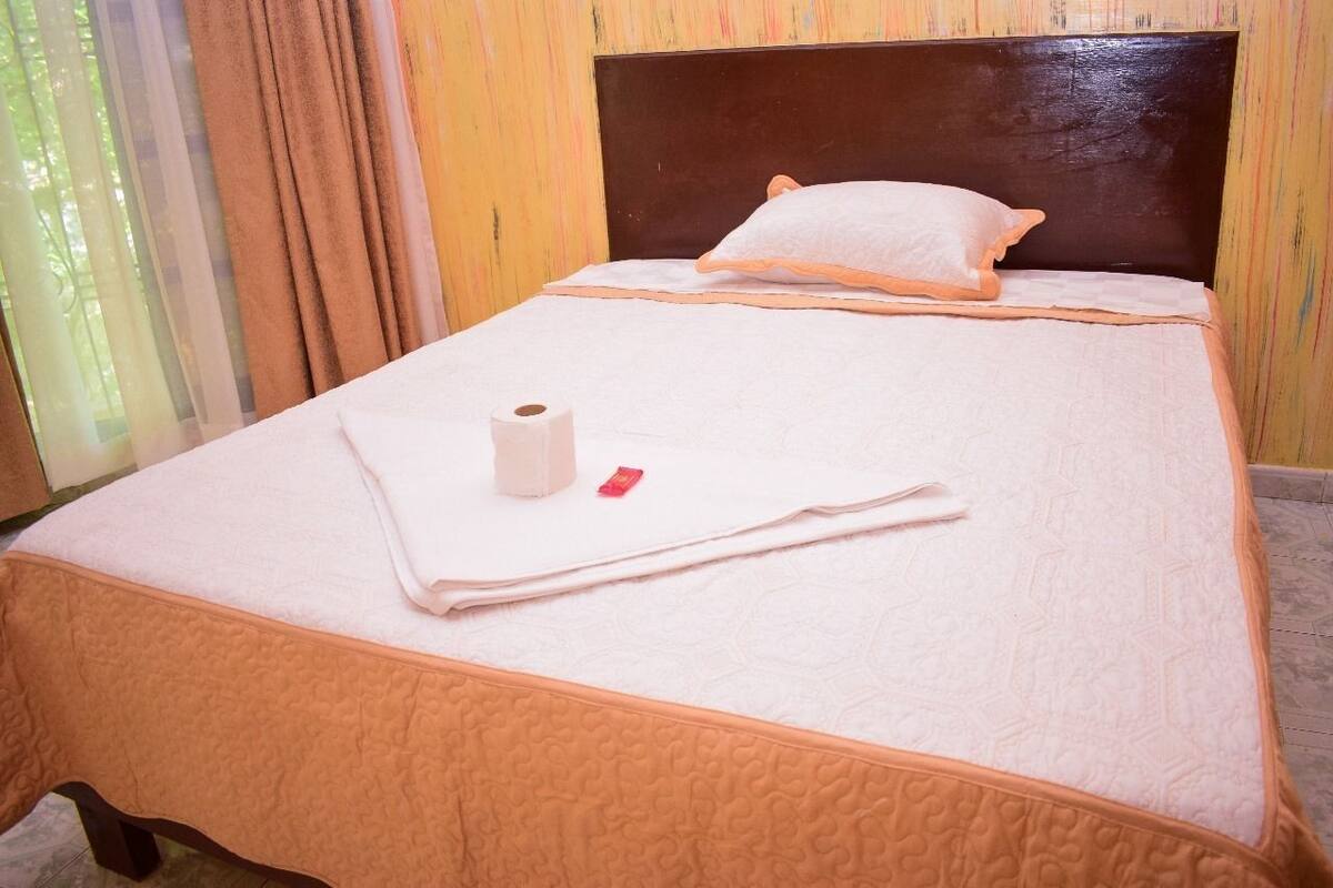 Cama bem arrumada com travesseiro, toalha enrolada e um pequeno objeto vermelho sobre a cama.