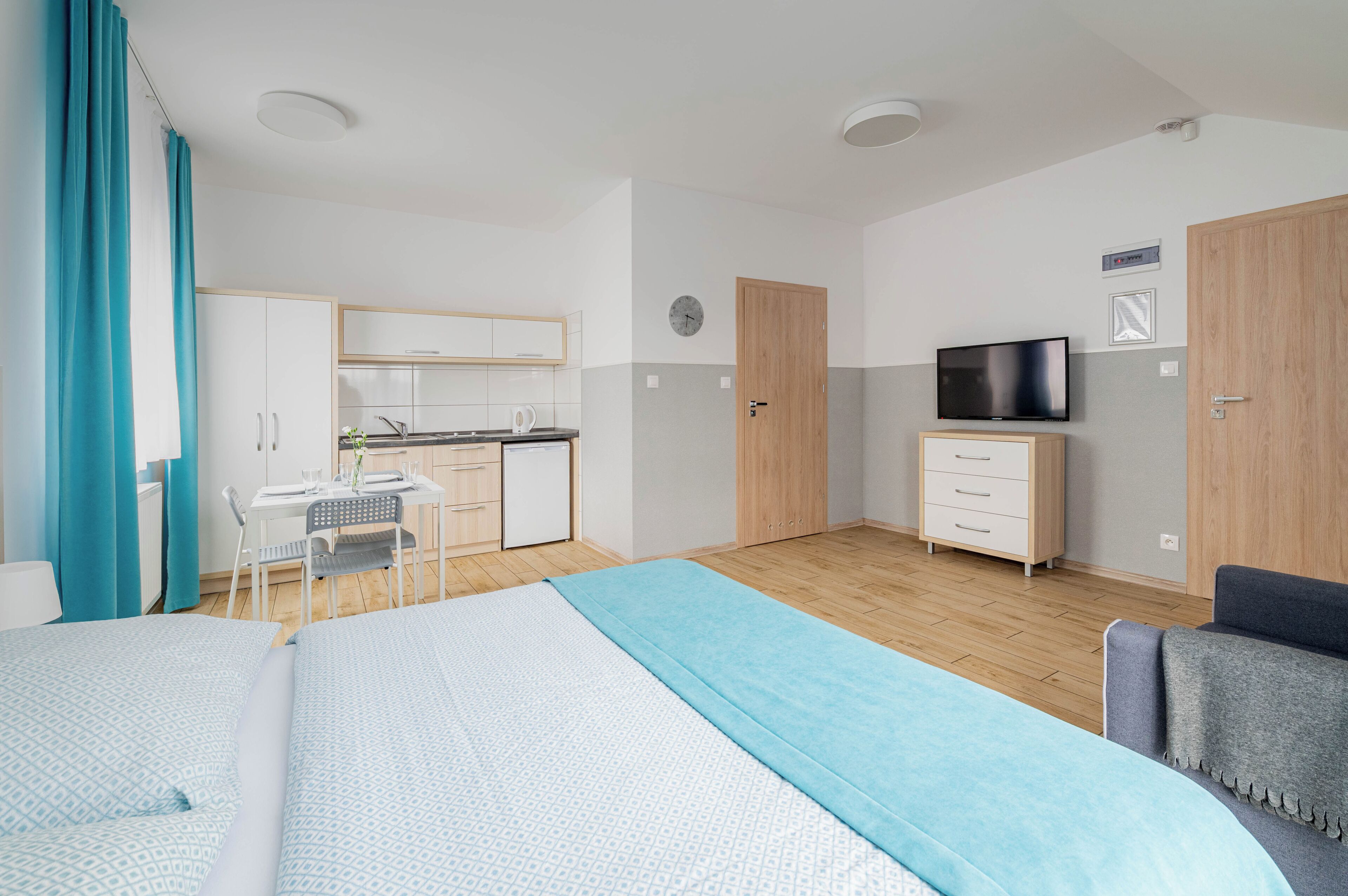 Quarto de hotel moderno com cama, escrivaninha, televisão e cozinha compacta.