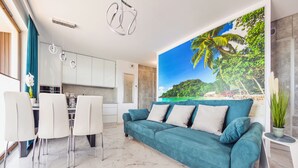 Uma sala moderna com um sofá azul-esverdeado, uma área de jantar com cadeiras brancas, uma cozinha com armários brancos e um grande mural de uma praia tropical na parede.