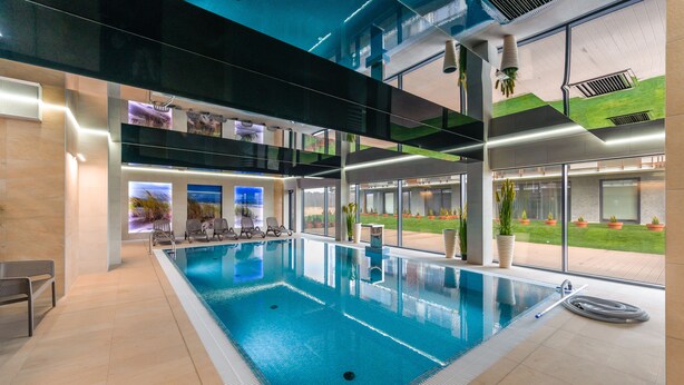 Piscina interior com água azul cristalina, rodeada por espreguiçadeiras e vista para um jardim ajardinado através de grandes janelas.