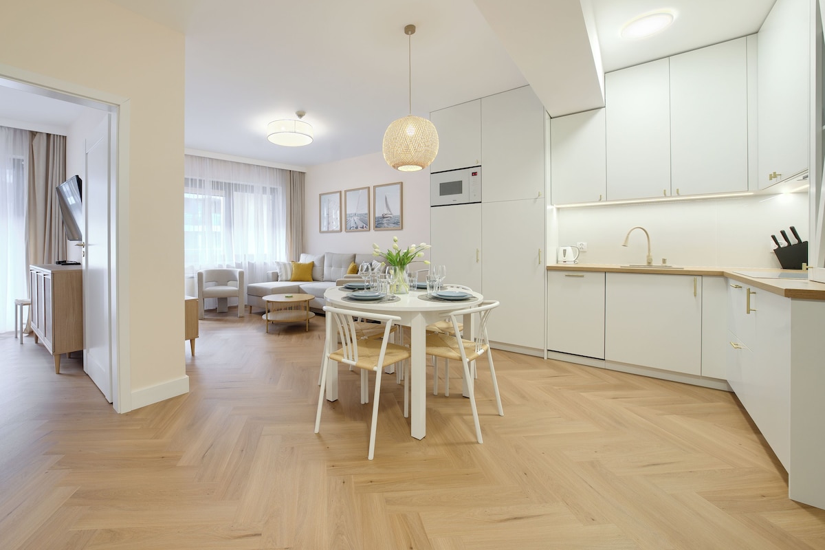 Cozinha moderna com armários brancos, área de refeições com mesa redonda e sala de estar com sofá e cortinas.