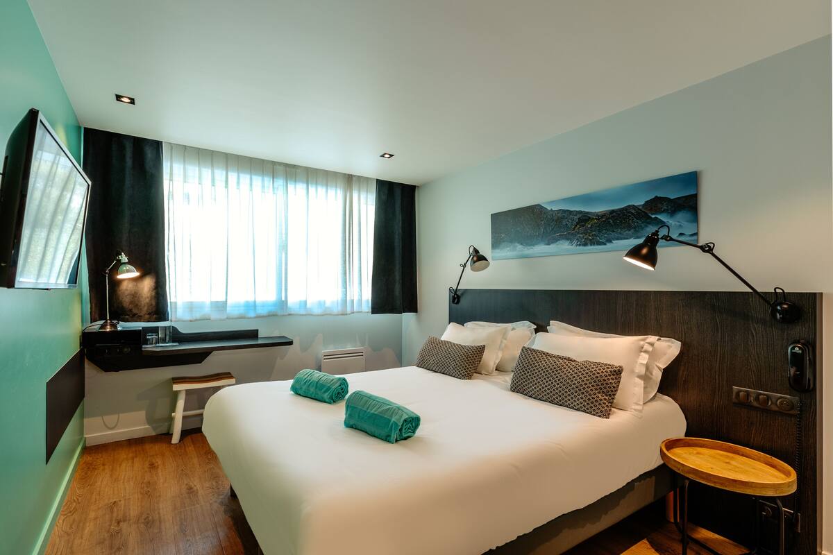 Quarto de hotel moderno com uma cama grande, uma escrivaninha com uma cadeira, um banquinho pequeno, uma luminária de mesa e um quadro na parede.