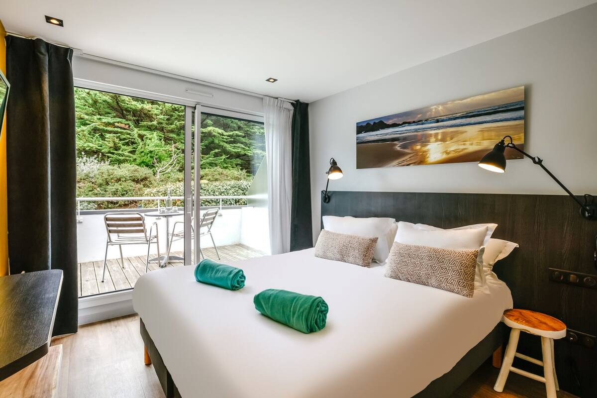 Quarto de hotel com uma cama grande, varanda com vista para árvores, um quadro na parede e uma escrivaninha com uma cadeira.