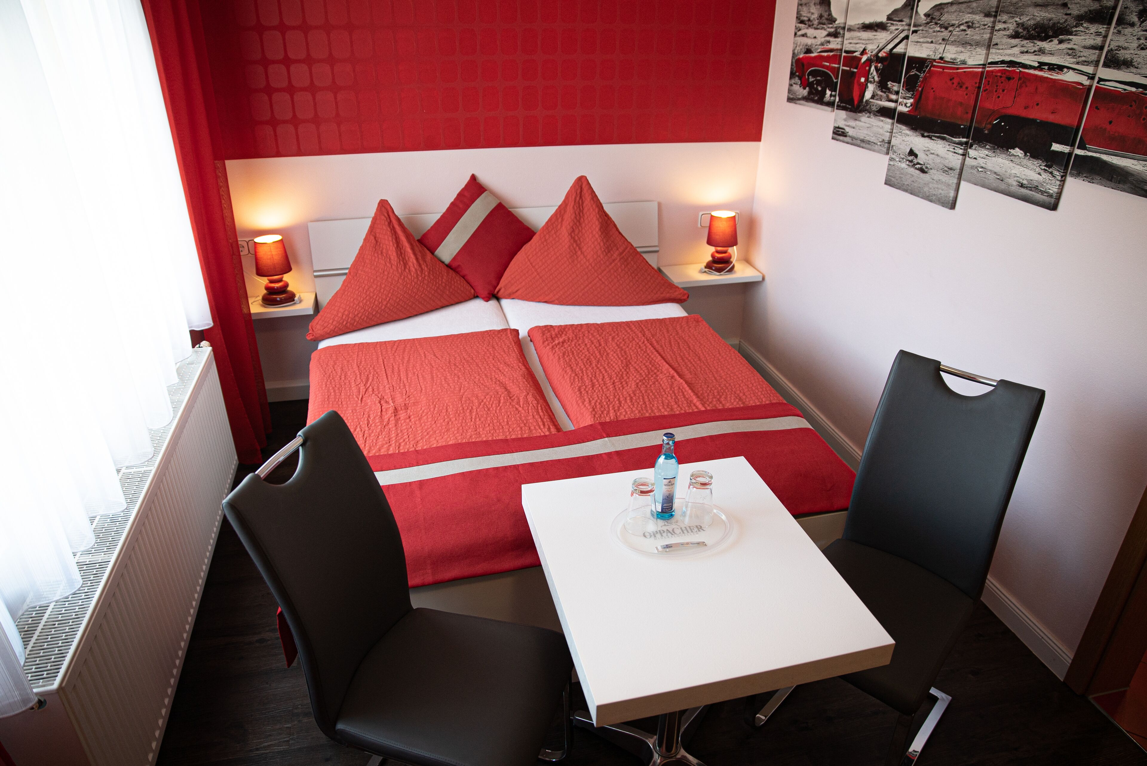 Quarto de hotel com uma cama, duas mesas de cabeceira com luminárias, uma mesinha com uma garrafa, duas cadeiras e um radiador.