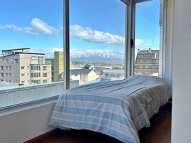 Cama em quarto com grandes janelas com vista para paisagem urbana e montanhas.