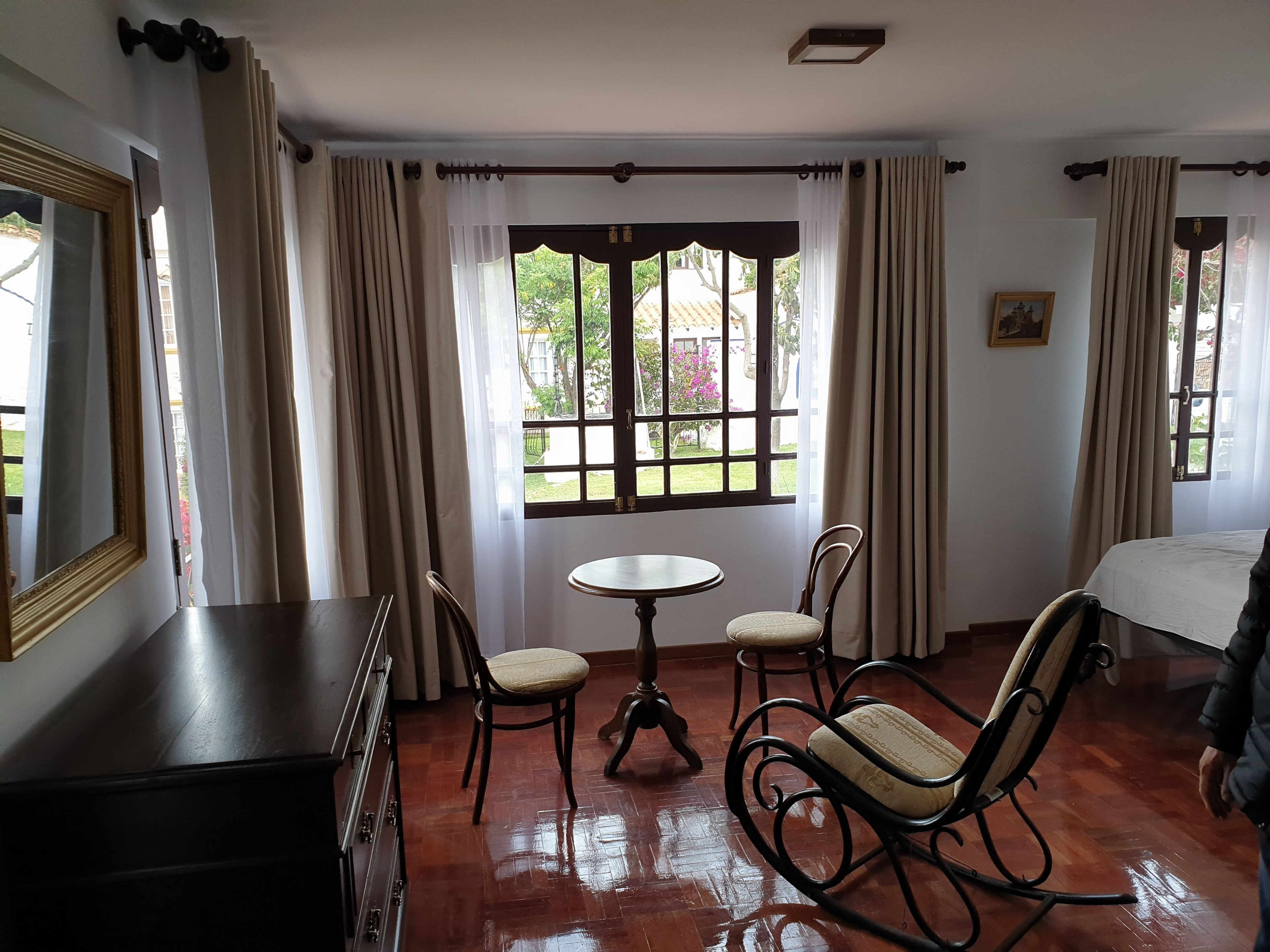Quarto com chão de madeira, mesa redonda com cadeiras, cama e janela com cortinas.