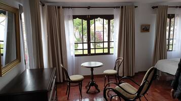 Quarto com chão de madeira, mesa redonda com cadeiras, cama e janela com cortinas.