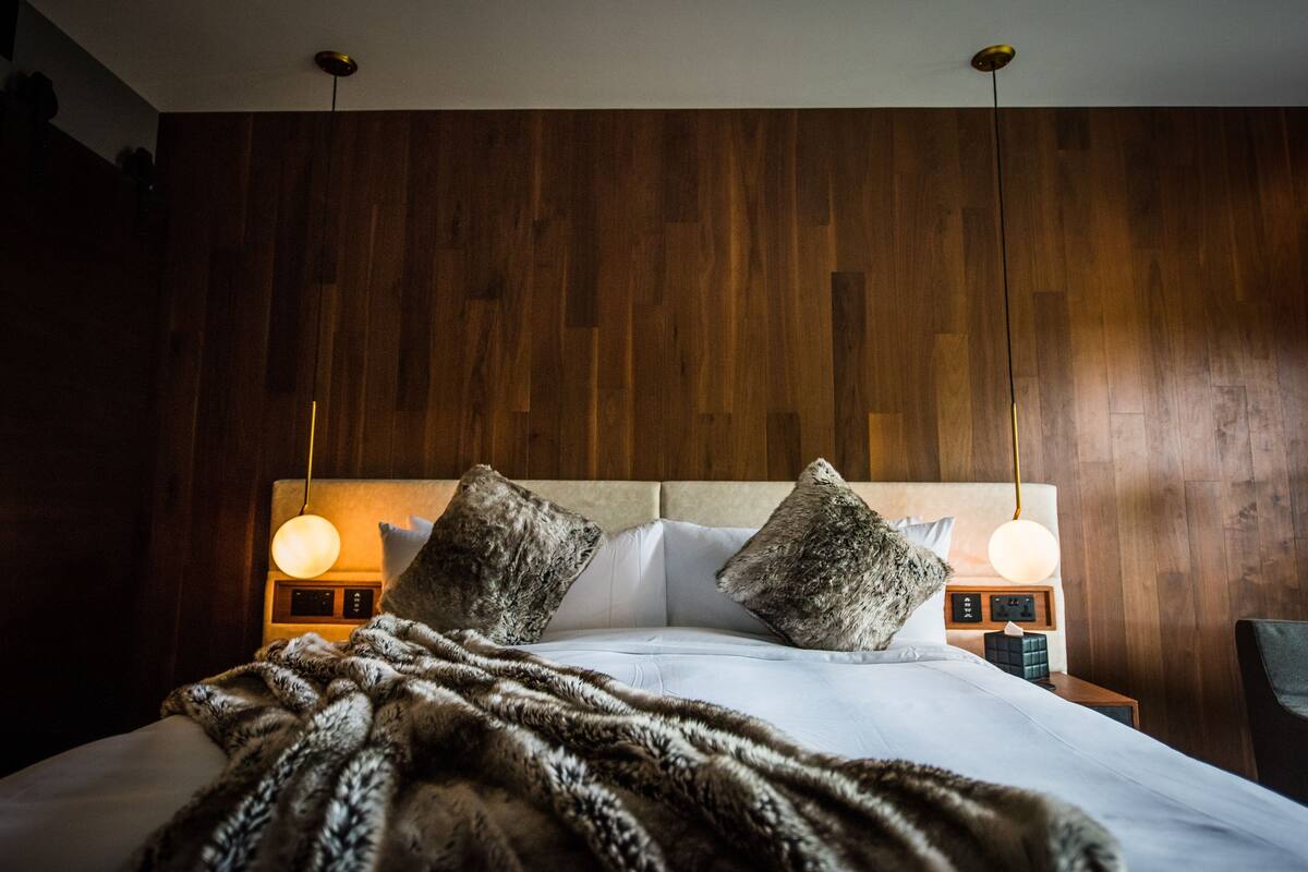 Cama bem arrumada com manta fofa, duas luminárias de cabeceira e cabeceira de madeira.