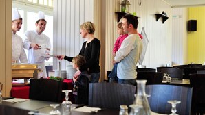 Restaurante com mesas preparadas para refeições, um garçom atendendo um cliente e chefs uniformizados.