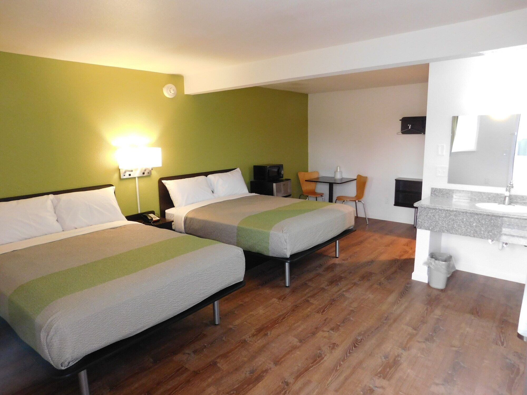 Quarto de hotel com duas camas, uma escrivaninha, uma cadeira, uma mesinha, um espelho e uma pia.