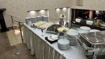 Disposição de buffet com diversos alimentos, utensílios, guarda-corpo de vidro e piso ladrilhado.
