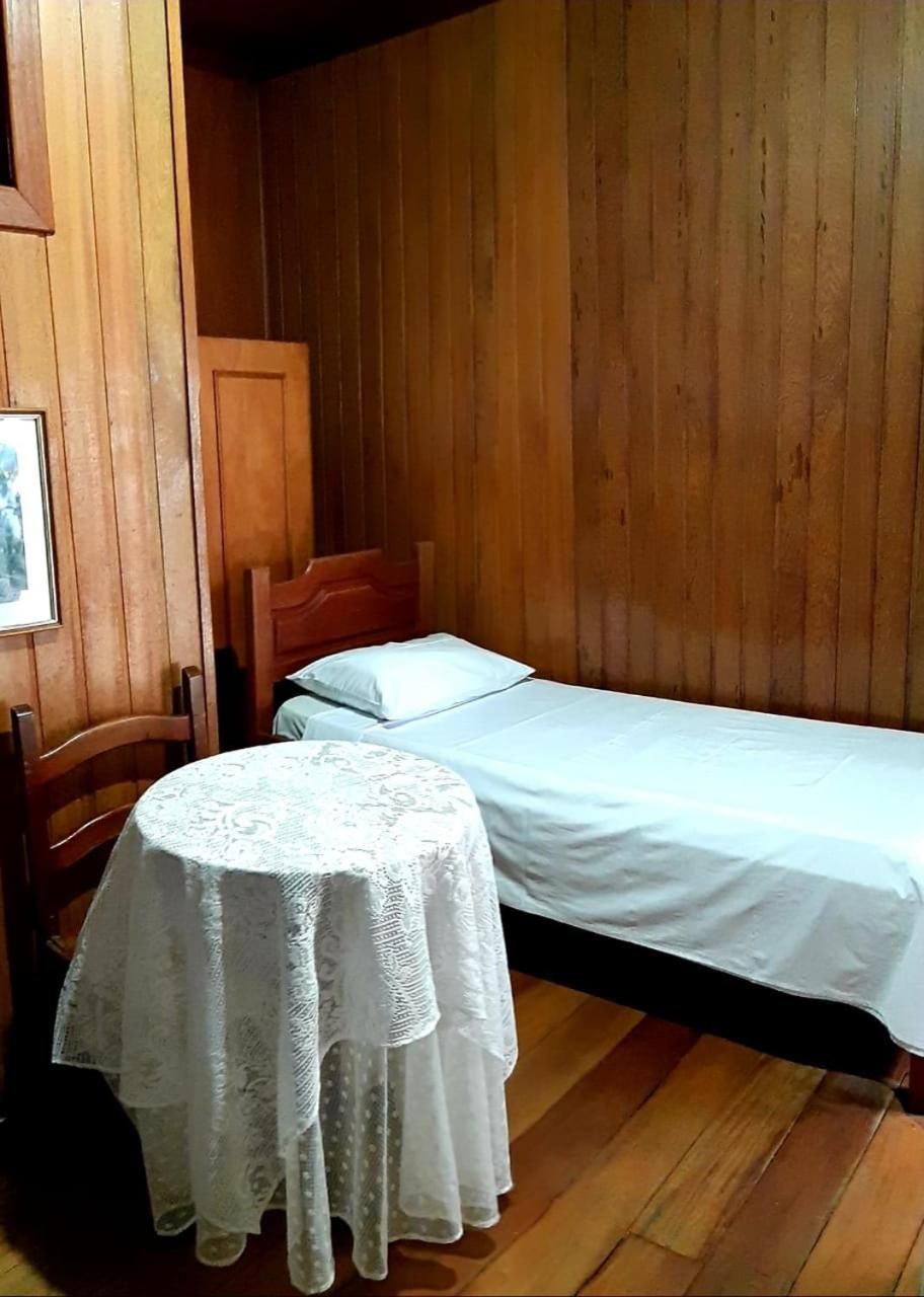 Quarto pequeno com uma cama de solteiro, cabeceira de madeira, mesa redonda com toalha de renda e chão de madeira.