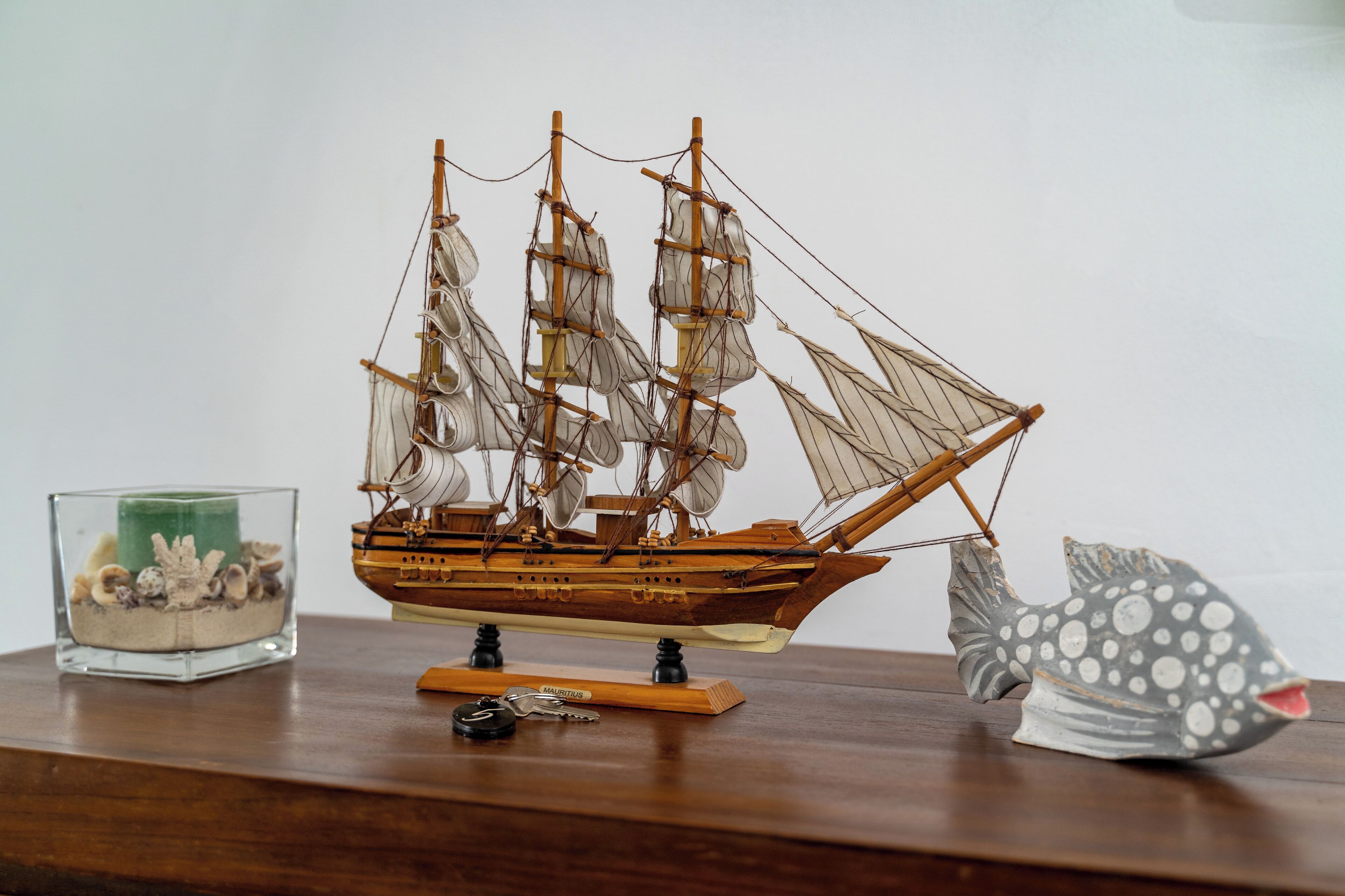 Um modelo de navio de madeira sobre uma superfície de madeira, um recipiente de vidro com conchas e um objeto em formato de peixe.