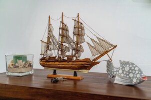 Um modelo de navio de madeira sobre uma superfície de madeira, um recipiente de vidro com conchas e um objeto em formato de peixe.