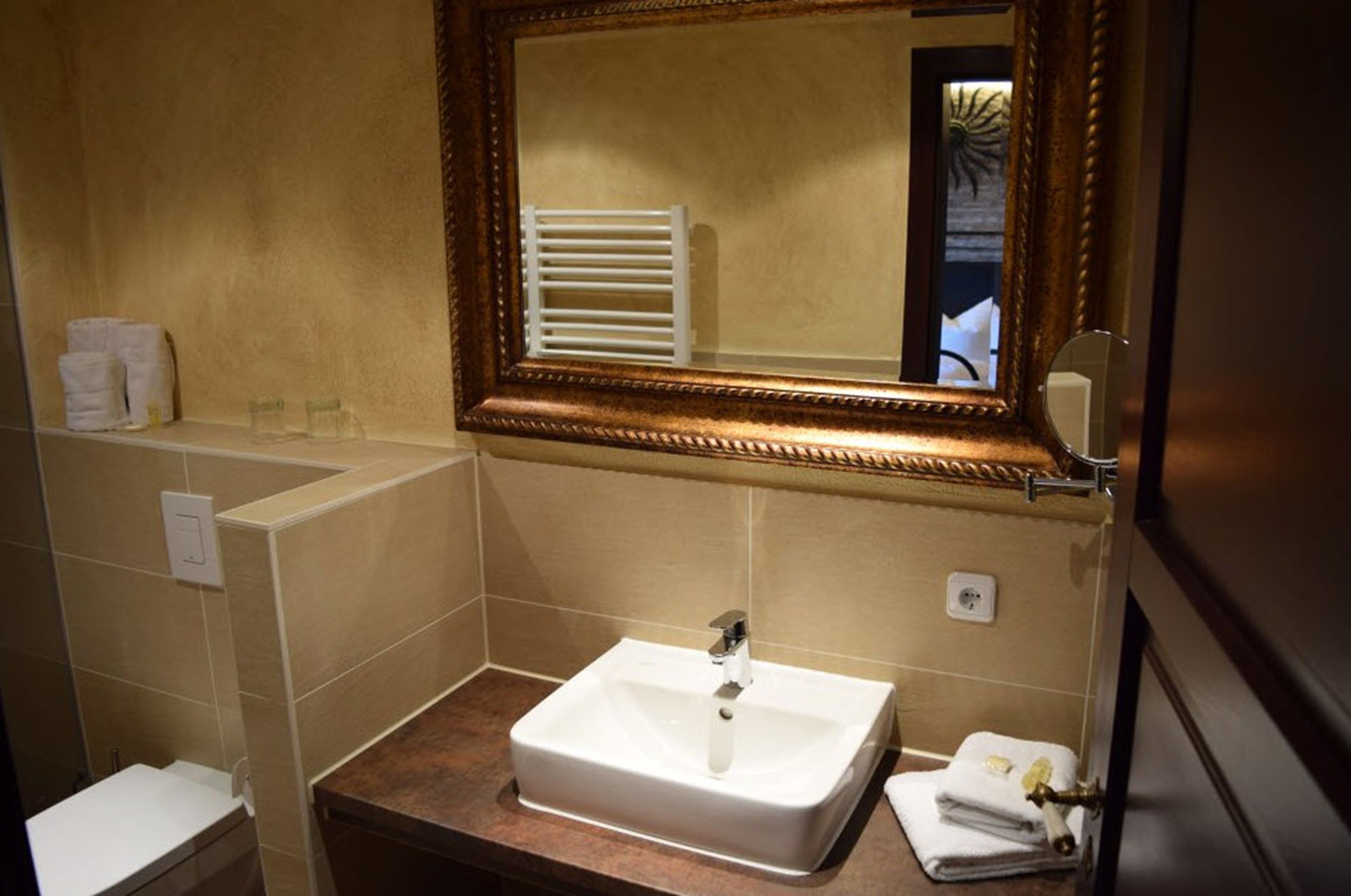 Casa de banho com lavatório branco, espelho com moldura dourada e um porta-toalhas.