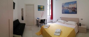 Quarto de hotel com cama, escrivaninha, cadeira, sofá, janela com cortinas vermelhas e obras de arte nas paredes.