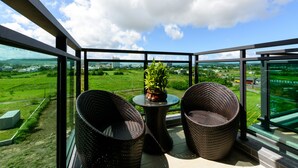 Uma varanda com cadeiras de vime e uma mesa, com vista para uma paisagem verde e uma área urbana.