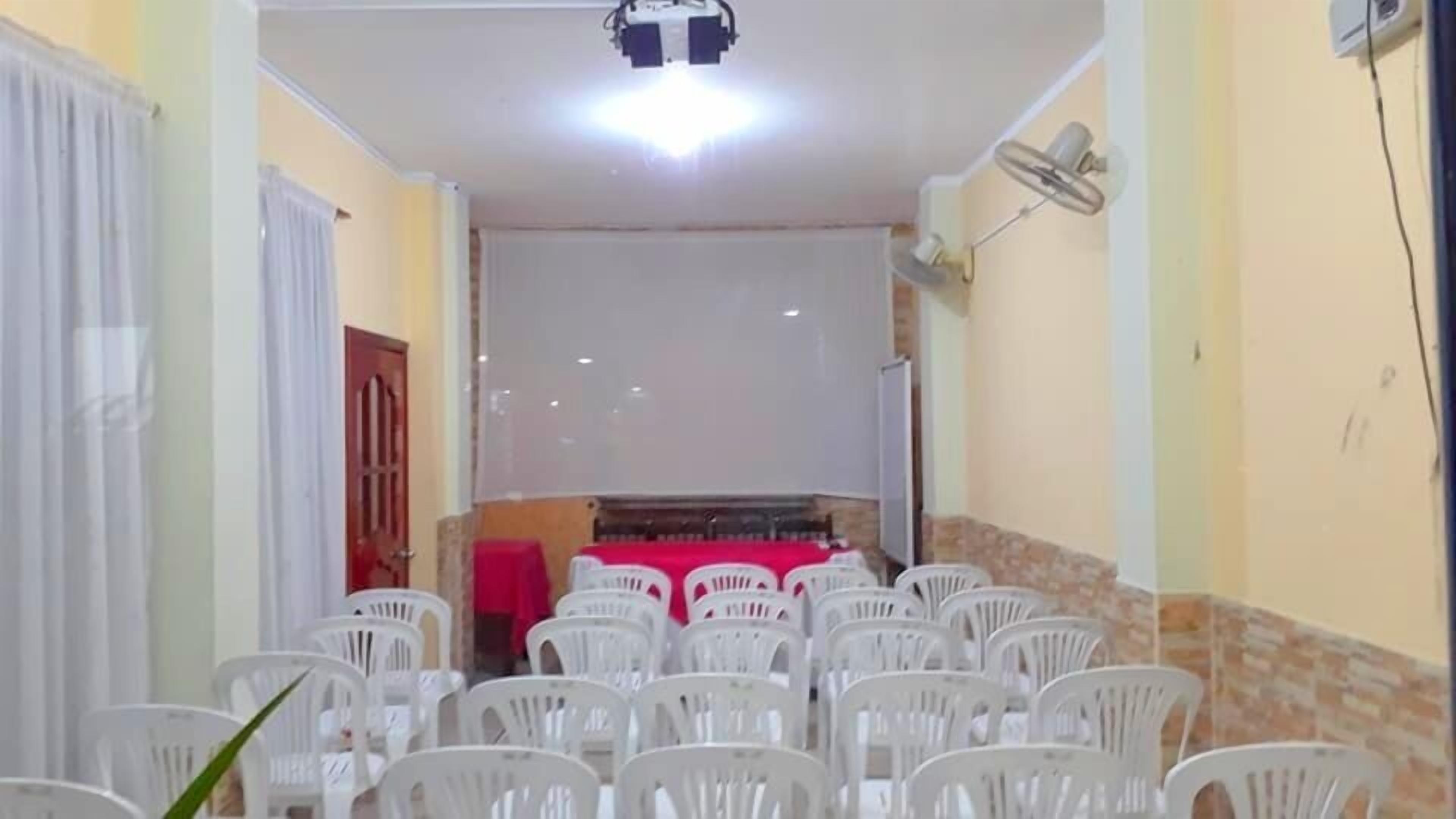 Uma sala de conferências com cadeiras de plástico branco dispostas em fileiras, uma mesa vermelha na frente e um projetor no teto.