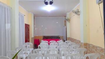 Uma sala de conferências com cadeiras de plástico branco dispostas em fileiras, uma mesa vermelha na frente e um projetor no teto.