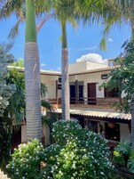 Um edifício de dois andares com fachada branca, varanda e jardim com plantas floridas brancas e palmeiras.