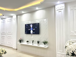 Uma área de recepção moderna com televisão de tela plana fixada em parede de mármore, balcão de recepção branco com plantas em vasos e molduras decorativas.