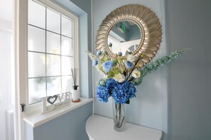 Casa de banho com toucador branco, espelho emoldurado a dourado e um vaso com flores azuis e brancas.